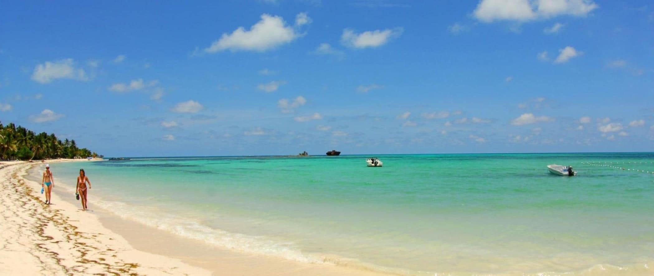 Uma praia com águas turquesa cristalinas, areal arenoso e alguns barcos ao longe.