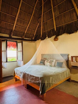 Um quarto com cama, janela com cortinas, telhado de palha e mesa de cabeceira.