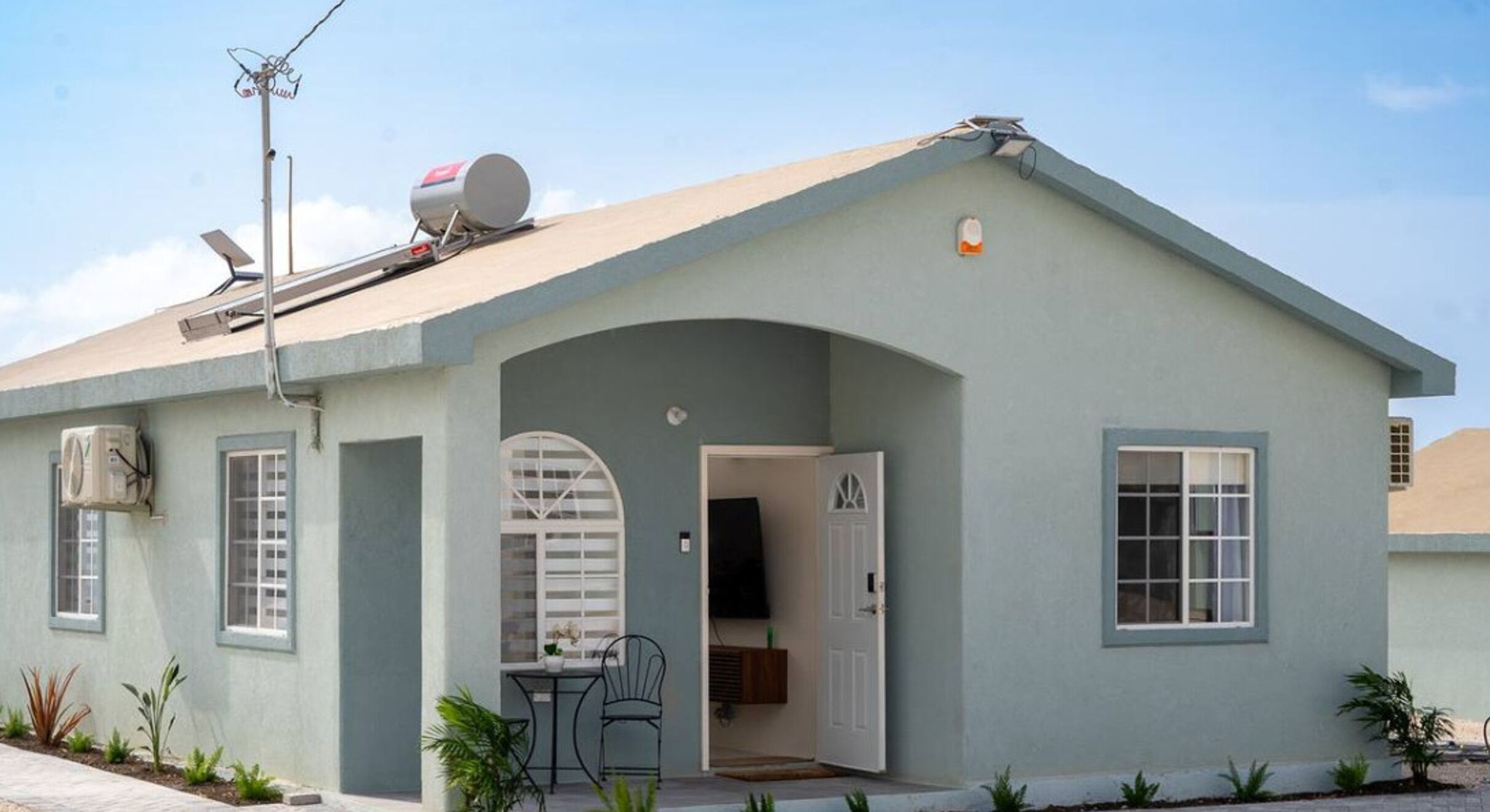 Uma casa térrea com telhado inclinado, varanda coberta com mesa e cadeiras, e antena parabólica no telhado.