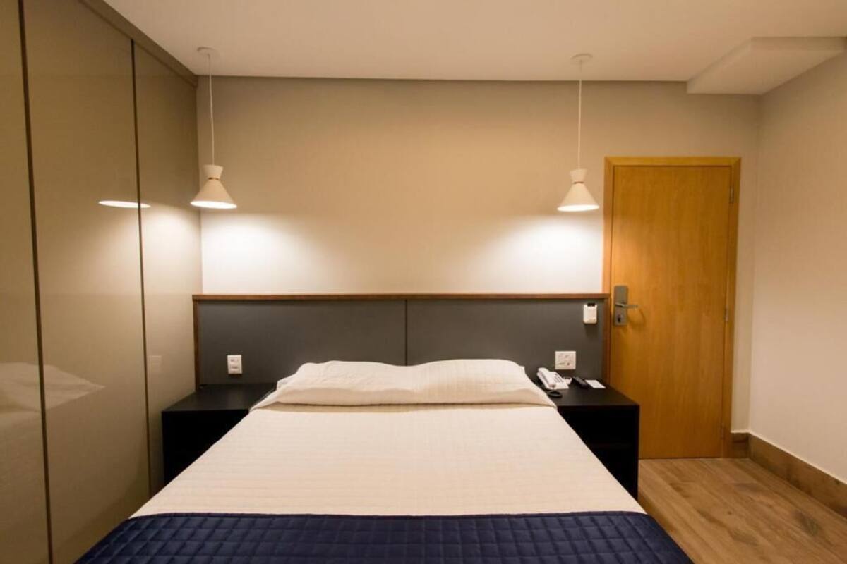 Quarto de hotel moderno com cama, mesas de cabeceira, porta de madeira e iluminação pendente.