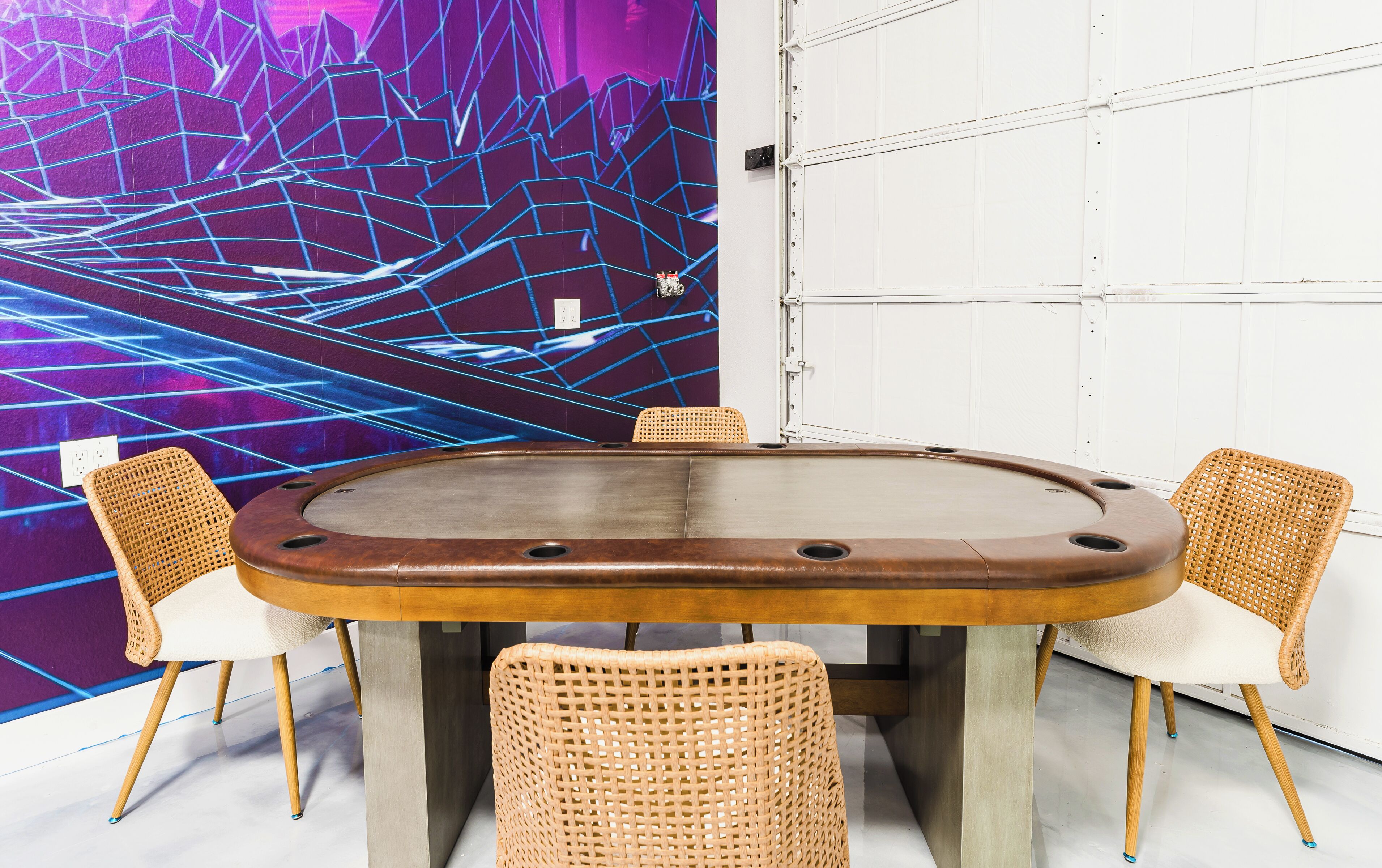 Uma mesa de bilhar com quatro cadeiras de vime, uma porta de garagem branca e um mural abstrato roxo na parede.