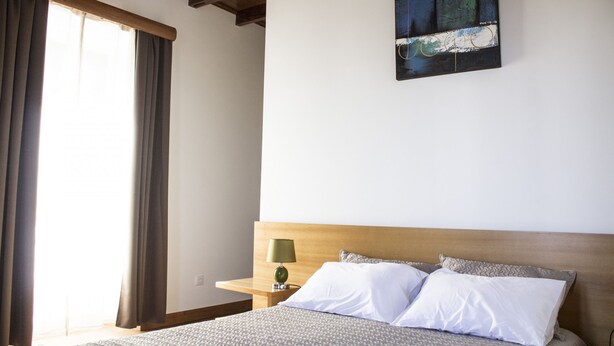 Quarto de hotel com cabeceira de madeira, cama com roupa de cama estampada, mesa de cabeceira com abajur e um quadro na parede.