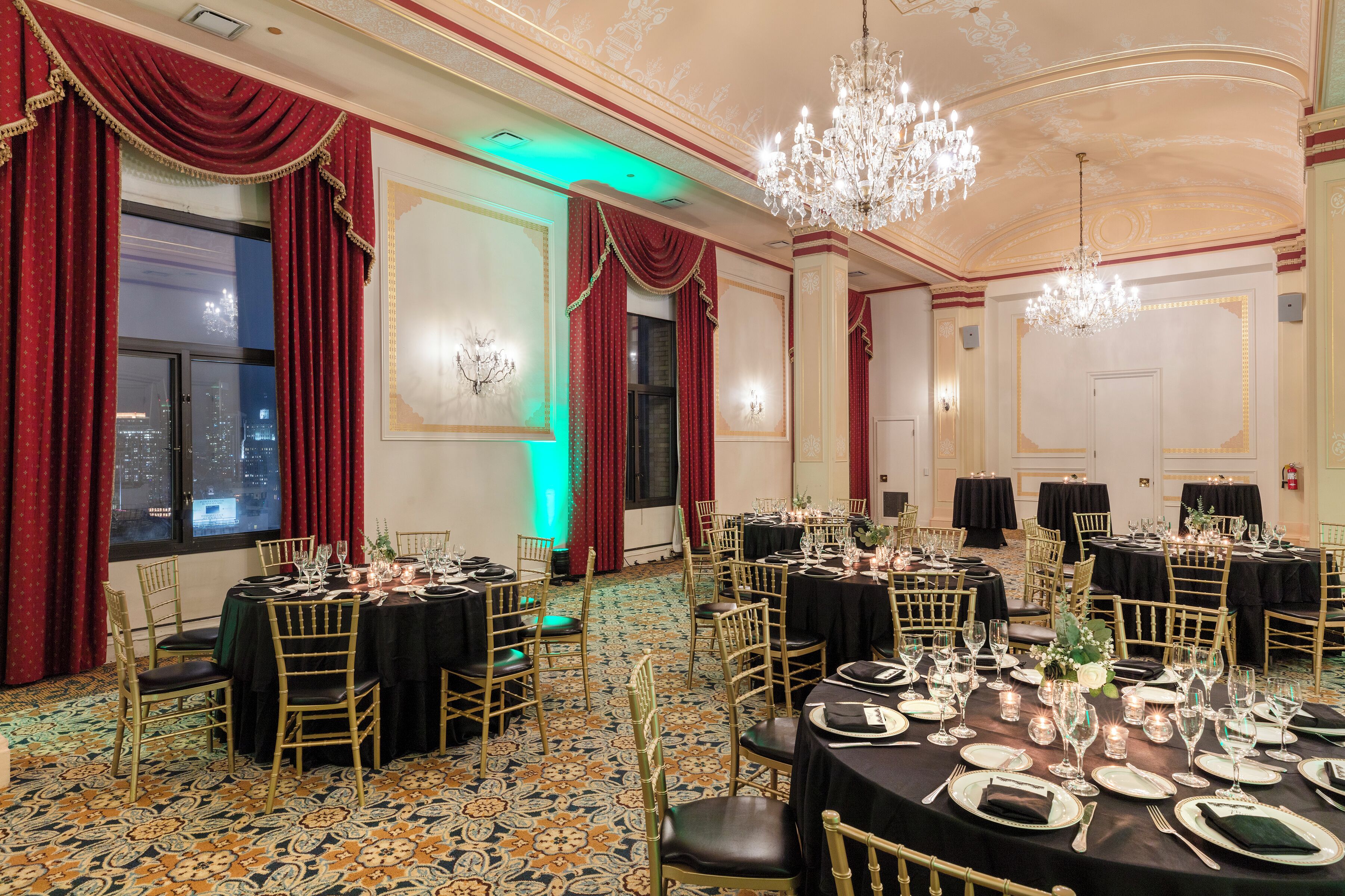 Salão de banquetes com mesas redondas, cadeiras douradas, lustres e cortinas vermelhas.