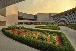 Um edifício moderno com fachada curva e um pátio ajardinado, com canteiros geométricos e uma escultura central.