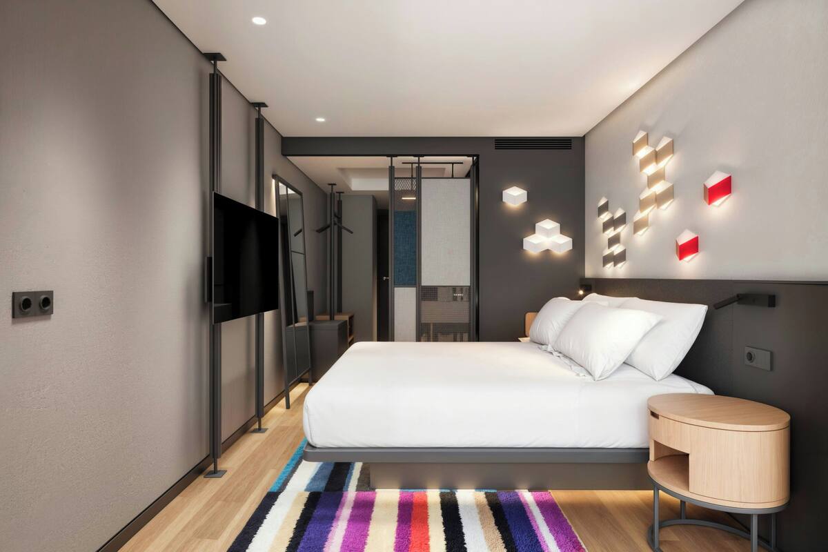 Quarto de hotel moderno com uma cama grande, uma mesinha de cabeceira, uma TV e um lustre de parede diferenciado.