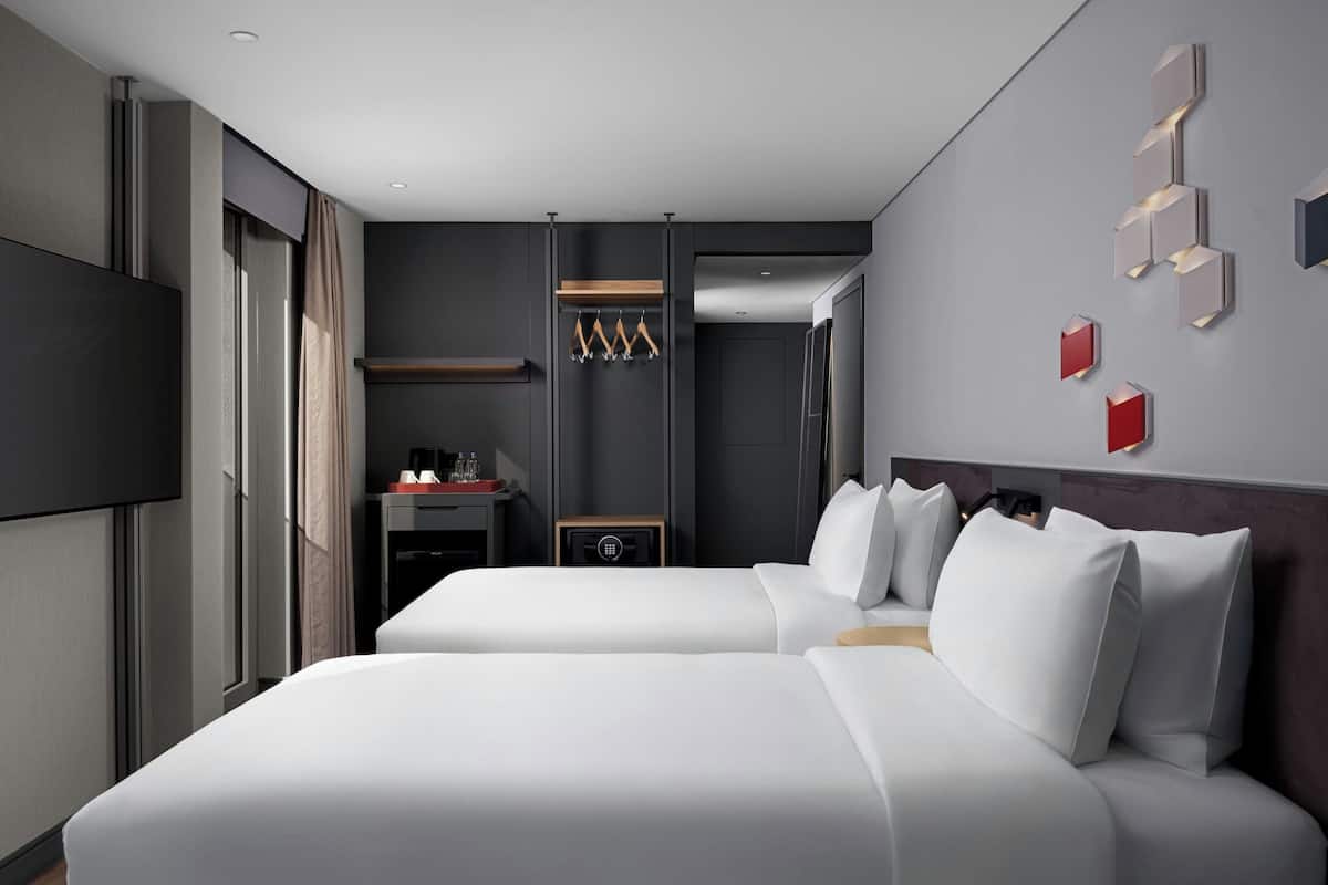 Quarto de hotel moderno com duas camas, uma TV de tela plana, uma pequena mesa de trabalho com cadeira e um porta-roupas.