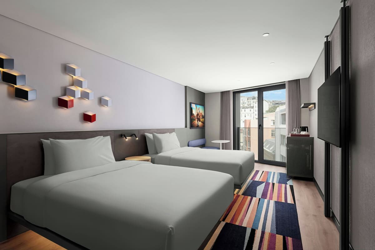 Quarto de hotel com duas camas, uma mesinha, uma televisão e uma janela com vista para a cidade.