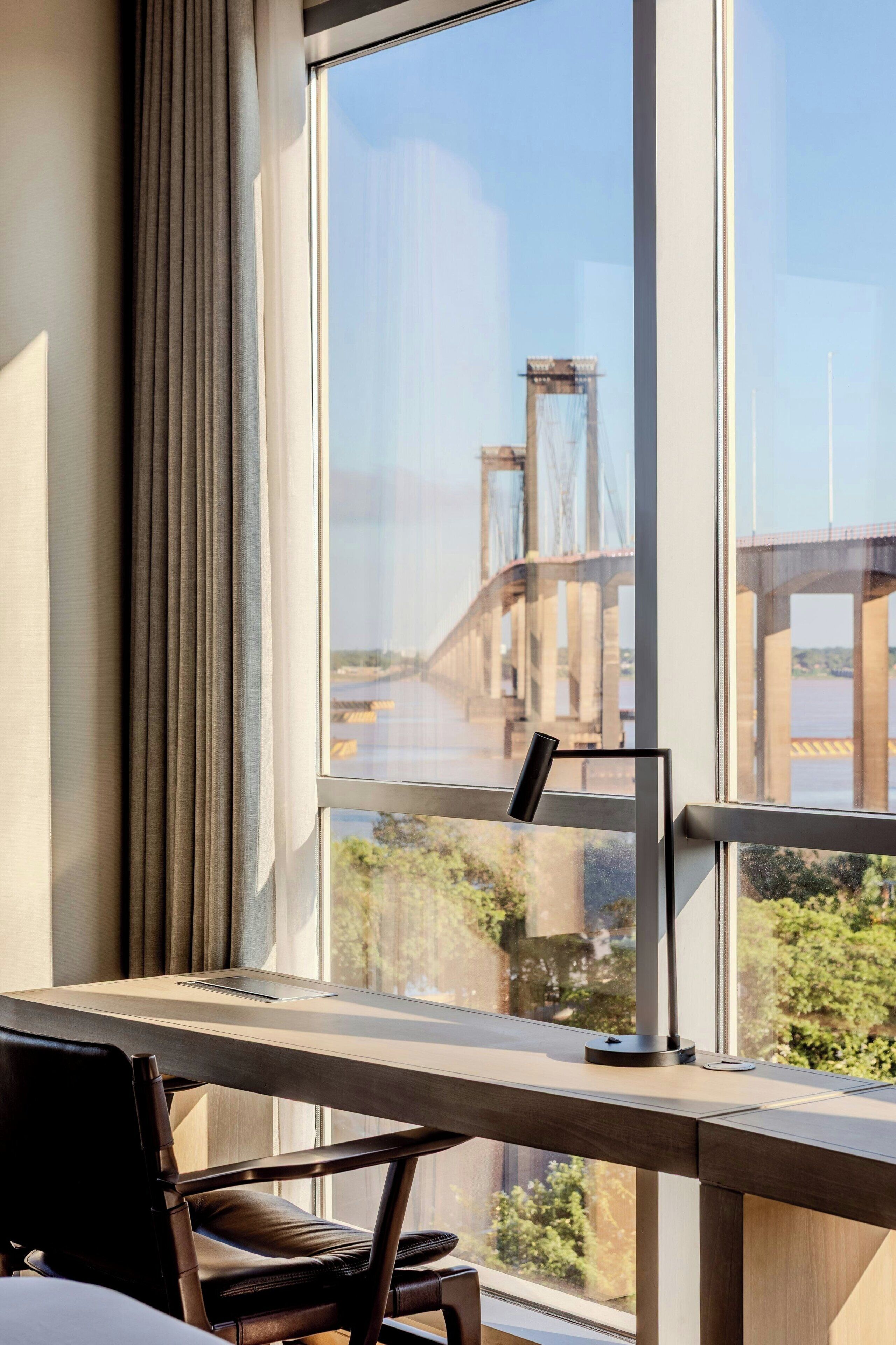 Quarto de hotel com uma grande janela, uma escrivaninha, uma cadeira e vista para uma ponte.