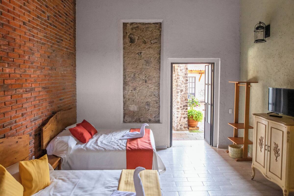Quarto de hotel com duas camas, parede de tijolos, ventilador de teto, armário de madeira e uma televisão.