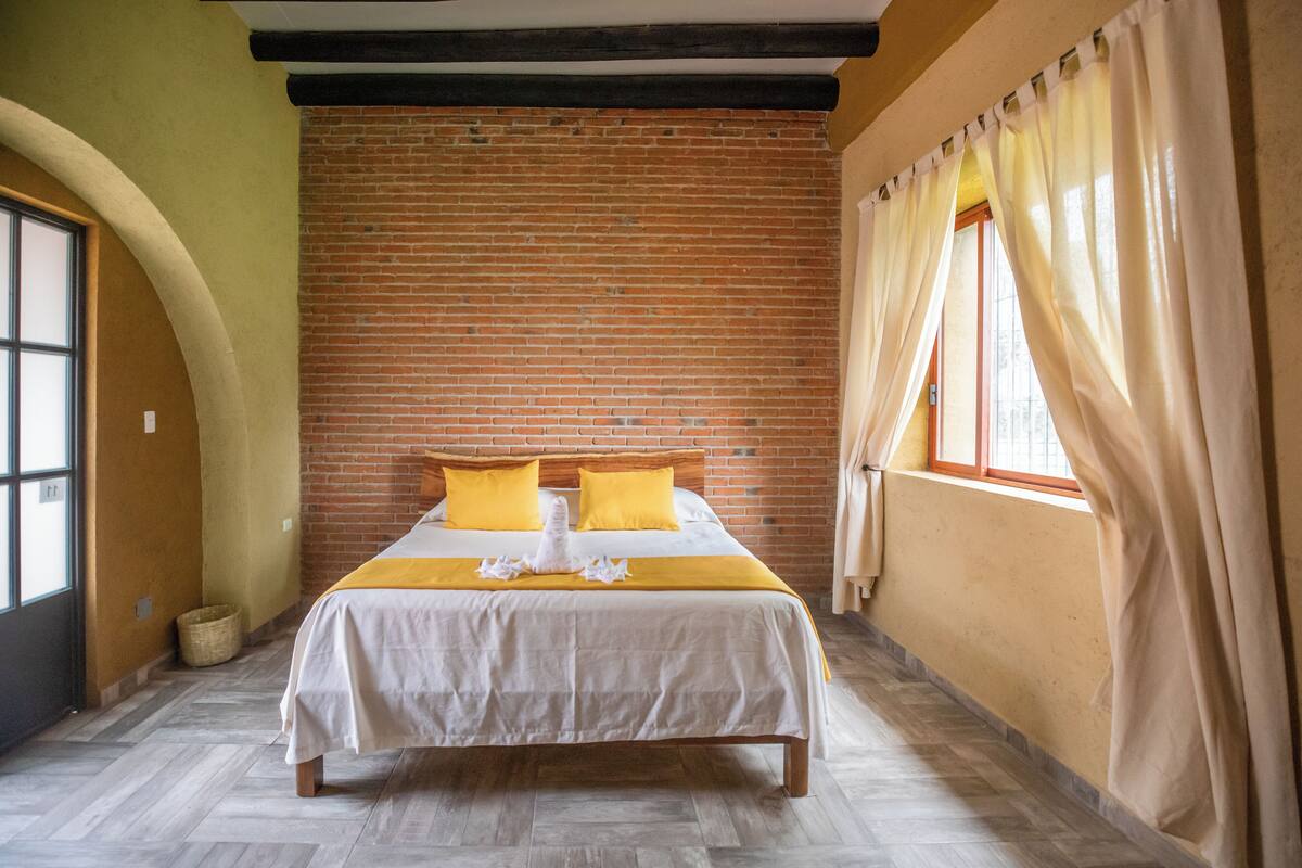 Quarto de hotel com parede de tijolos, cama com roupa de cama branca e amarela, e janela com cortinas.