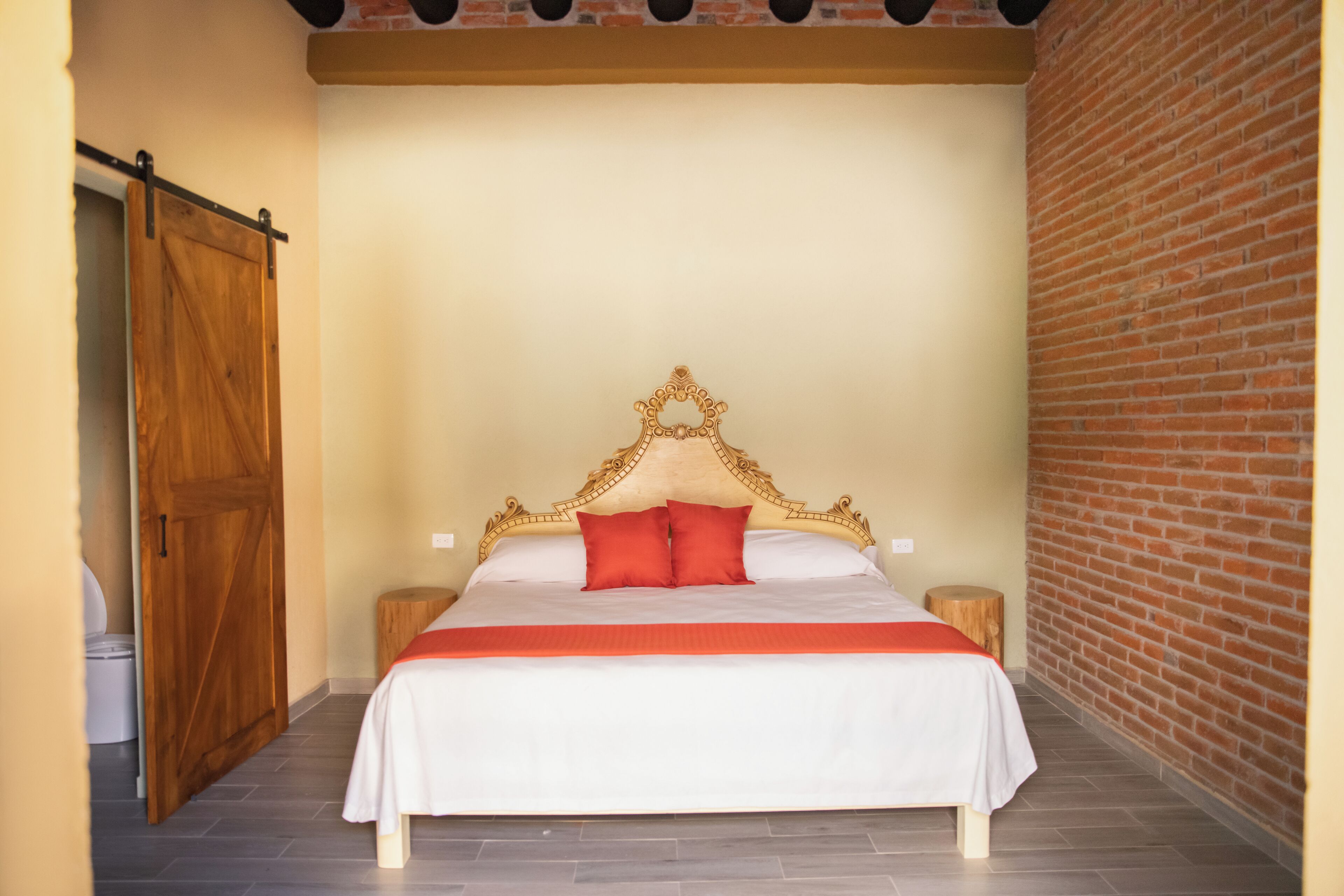 Quarto com uma cama grande, cabeceira de madeira, duas almofadas vermelhas, um edredom branco e um cobertor vermelho. Há também um banquinho de madeira e uma parede de tijolos à vista.