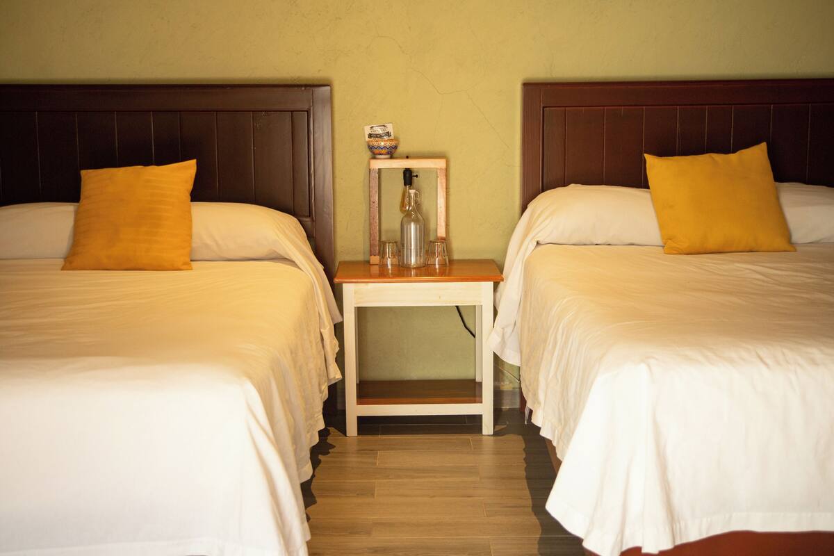 Quarto de hotel com duas camas, cada uma com roupa de cama branca e um único travesseiro amarelo. Uma pequena mesa de cabeceira de madeira com uma garrafa de vidro e uma tigela está colocada entre as camas.