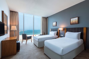 Quarto de hotel com duas camas, uma escrivaninha, uma cadeira e uma janela grande com vista para o oceano.