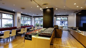 Um restaurante com um balcão comprido exibindo diversos pratos, uma área de estar com mesas e cadeiras, e grandes janelas que proporcionam luz natural.