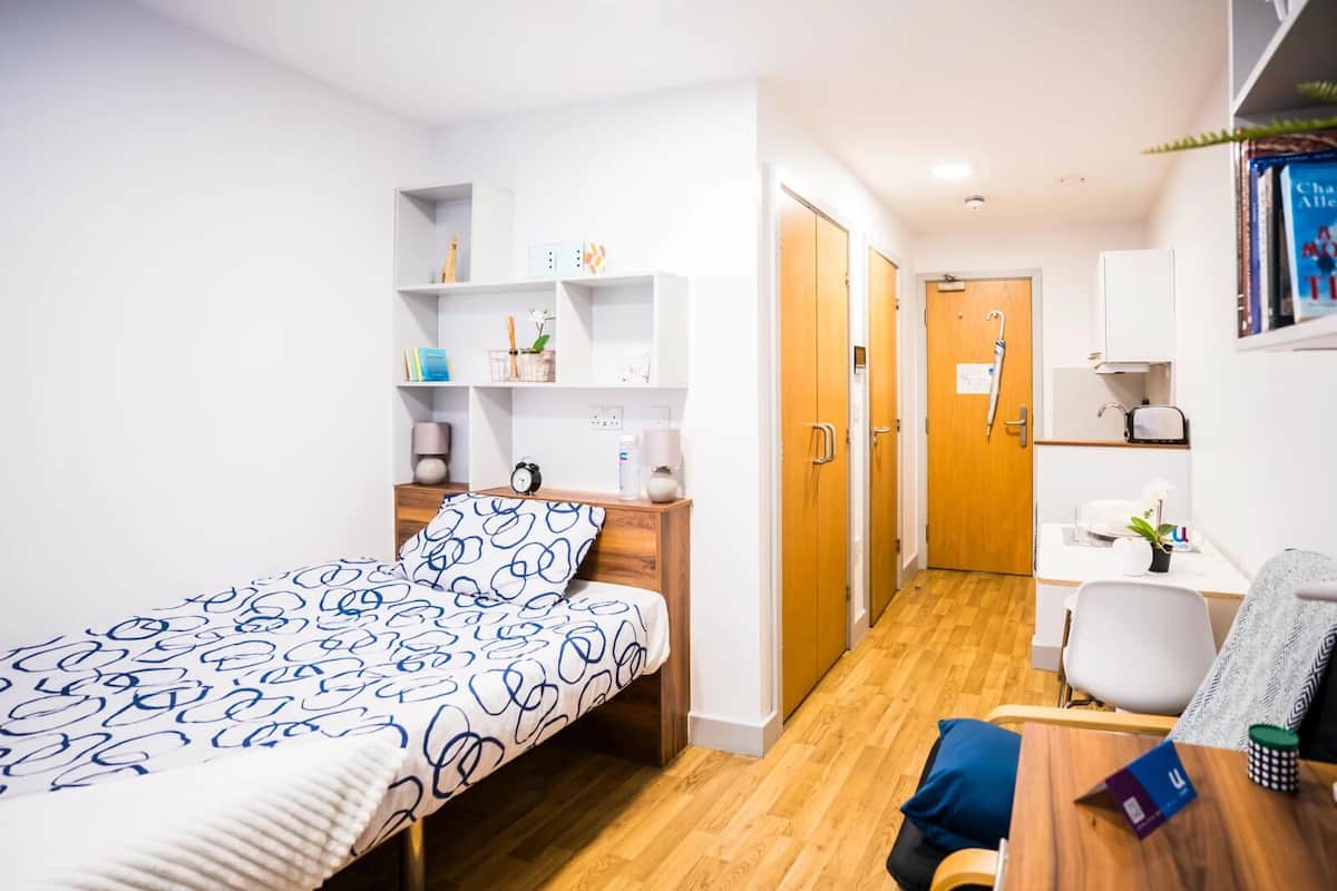 Quarto de hotel pequeno com uma cama de solteiro, uma escrivaninha, uma cadeira, um guarda-roupa e um banheiro.