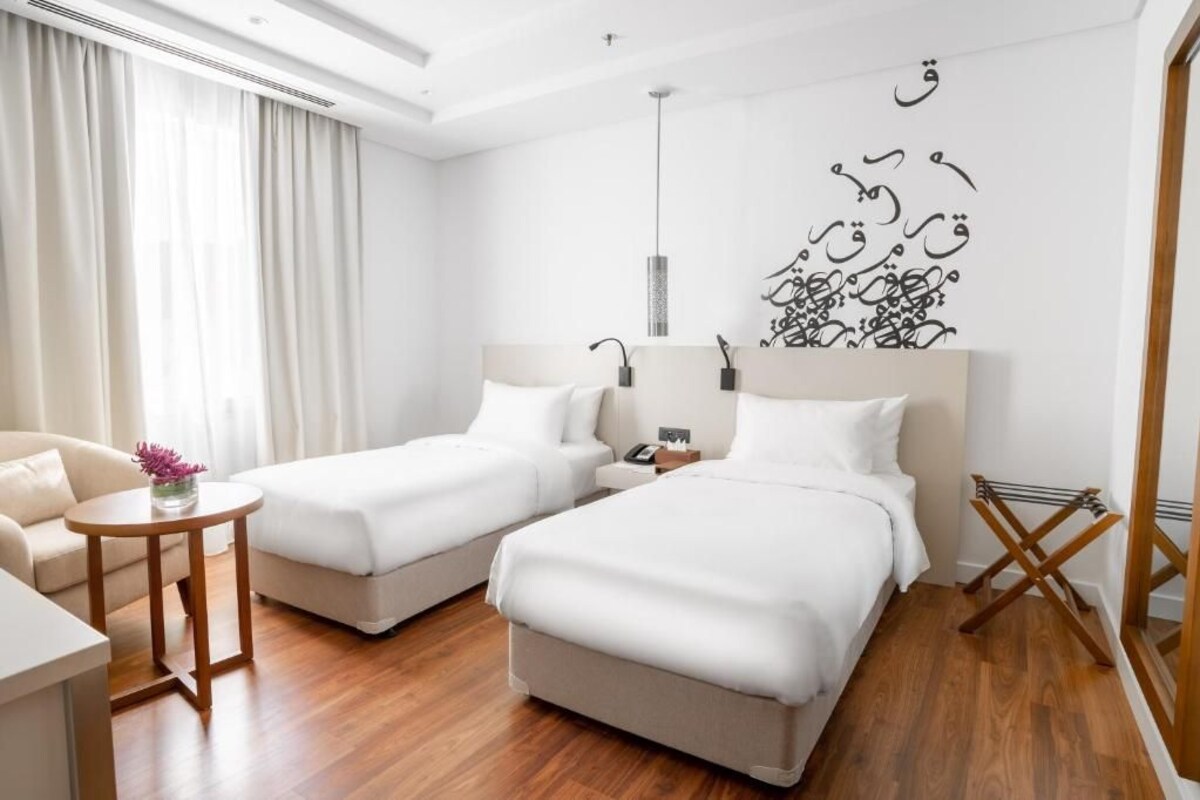 Quarto de hotel com duas camas, uma mesinha com um vaso, um sofá e uma obra de arte na parede.
