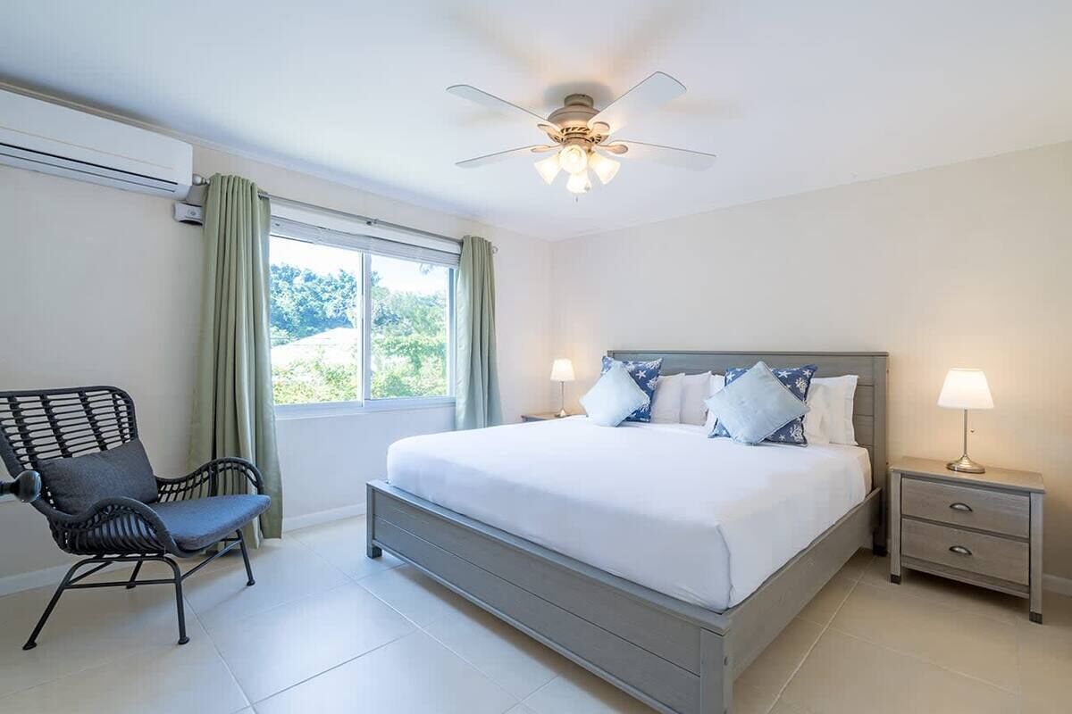 Quarto com cama, cadeira, mesa de cabeceira, ventoinha no teto e janela.