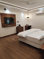Quarto moderno com televisão de tela plana fixada em um painel de madeira, mesa de cabeceira com luminária e cama com roupa de cama branca.