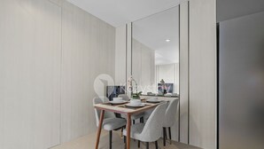 Uma sala de jantar moderna com mesa de madeira e cadeiras estofadas cinzentas, uma planta em vaso e uma cozinha ao fundo.