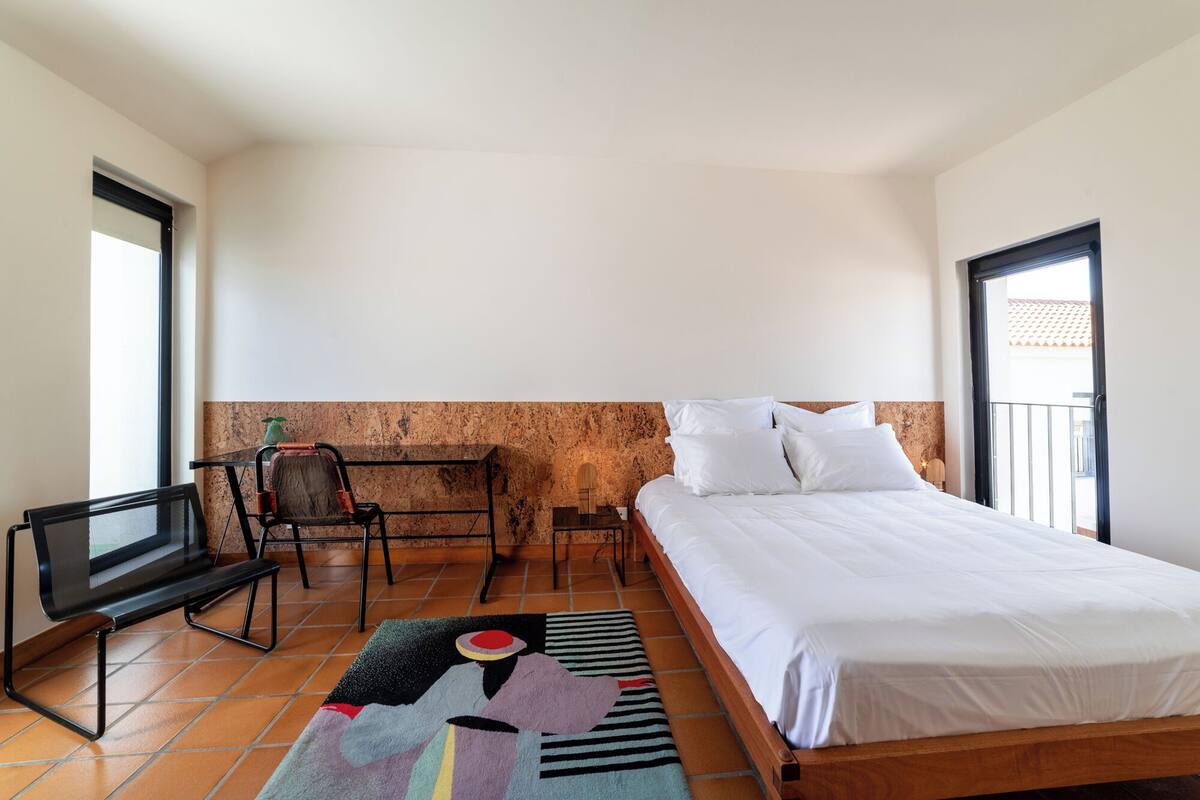 Quarto de hotel com uma cama, uma escrivaninha, uma cadeira e uma televisão.
