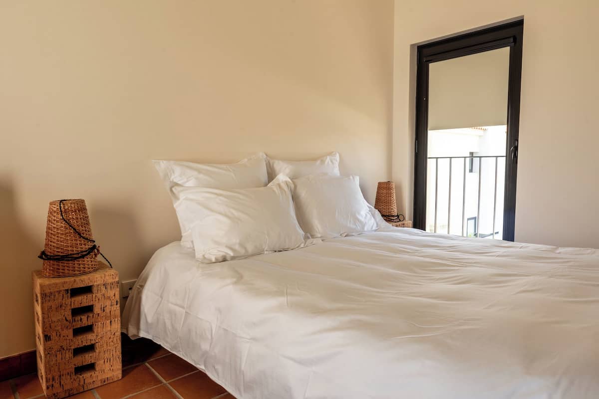 Uma cama com roupa de cama branca, um espelho grande na parede e um cesto de vime sobre um banquinho ao lado da cama.