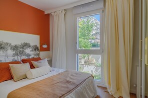 Quarto com cama, cortinas brancas, janela com vista para árvores e edifícios, e uma parede laranja.