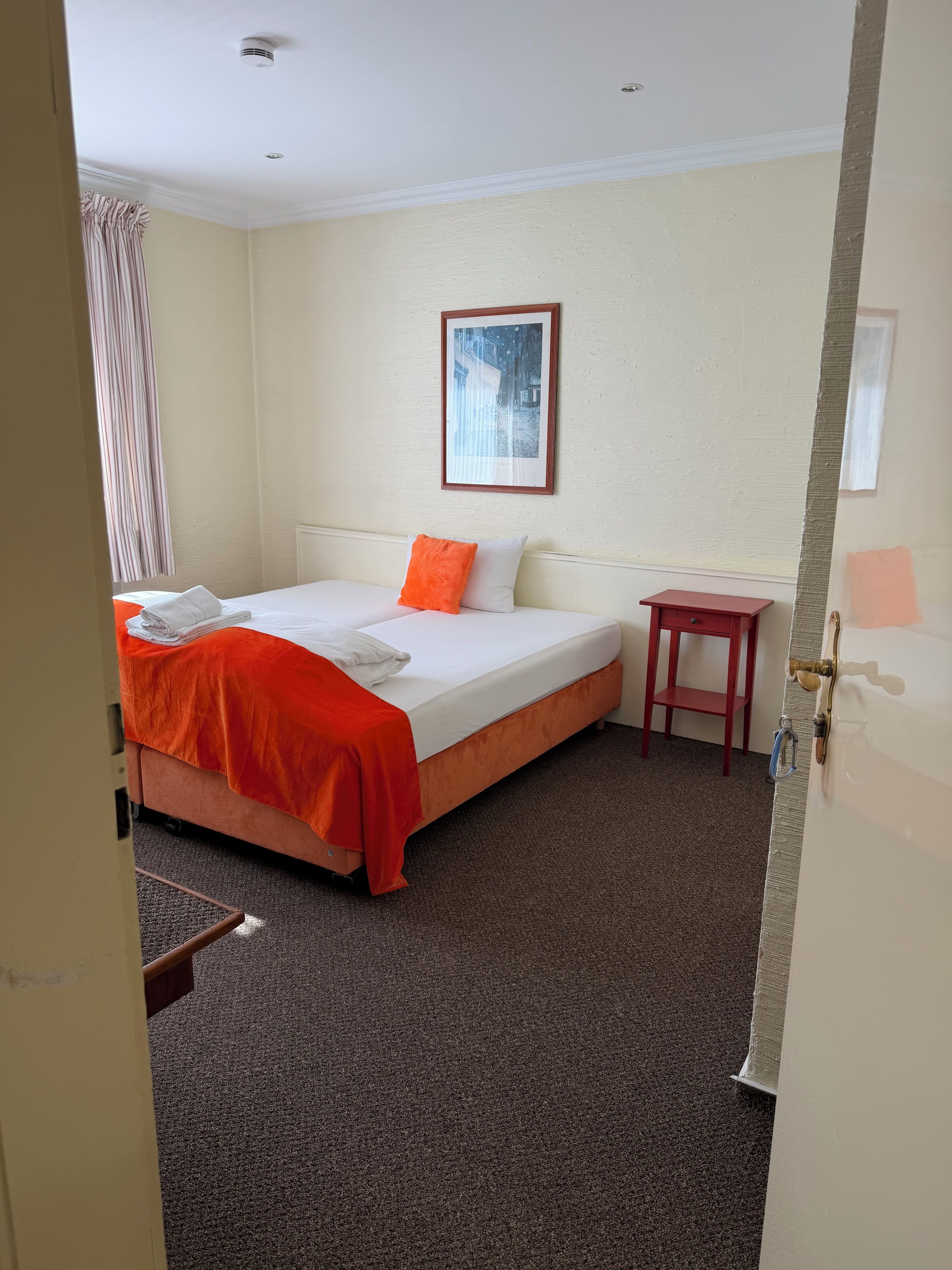 Quarto de hotel com cama, uma mesinha, uma cadeira vermelha, um quadro na parede e uma porta com tranca.