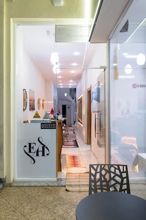 Uma área de recepção de hotel com porta de vidro, balcão de recepção, área de estar com cadeiras e parede com obras de arte e aviso de 'NÃO FUMAR'.