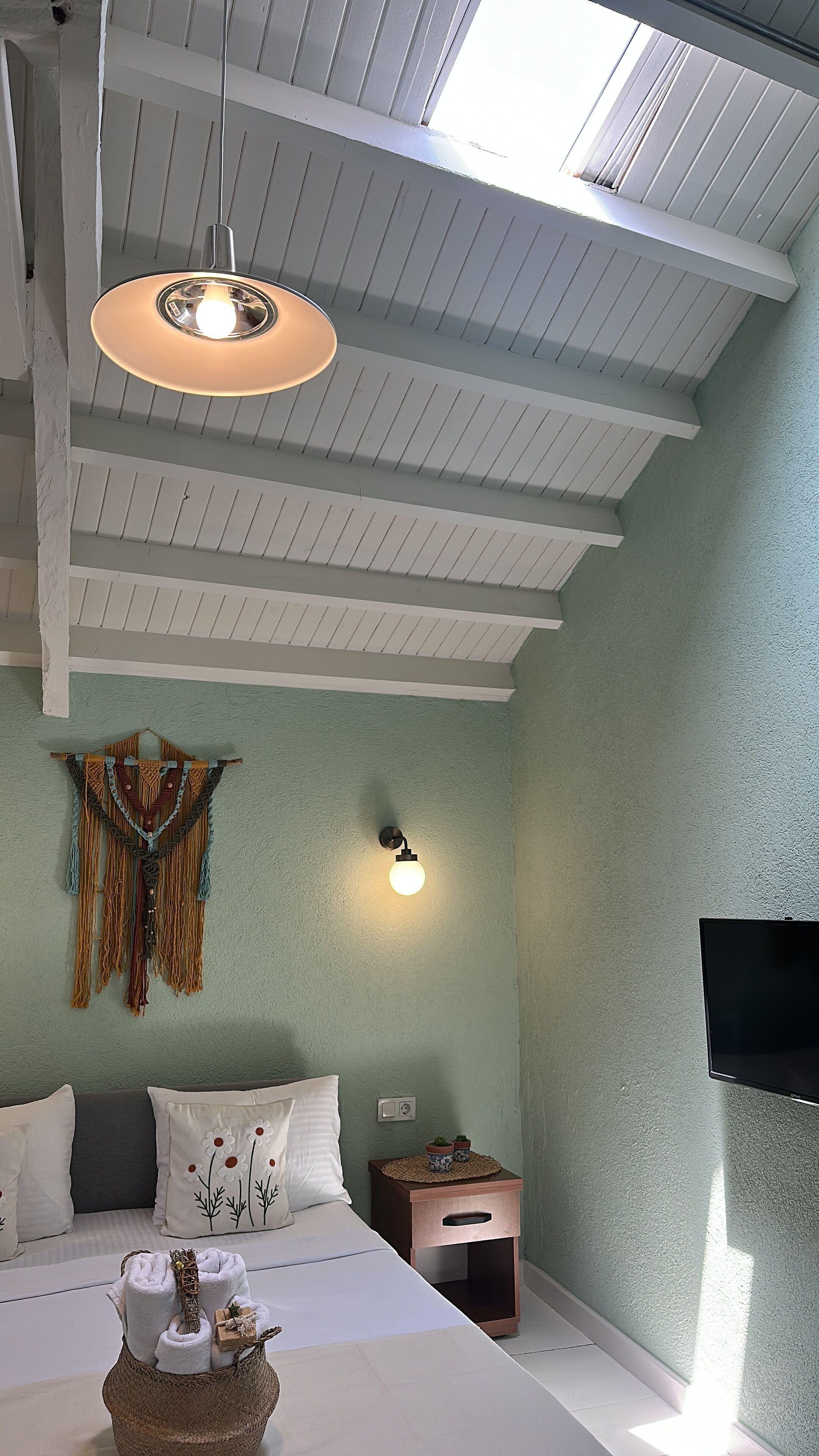 Quarto com cama, mesa de cabeceira, TV fixada na parede, ventoinha no teto e um objeto decorativo na parede.