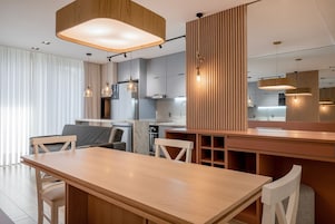 Uma área de refeições moderna com uma mesa e cadeiras de madeira, iluminação pendente e uma cozinha ao fundo.