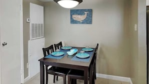 Uma área de refeições com uma mesa posta para quatro, cadeiras, luminária fixada na parede e uma obra de arte na parede.