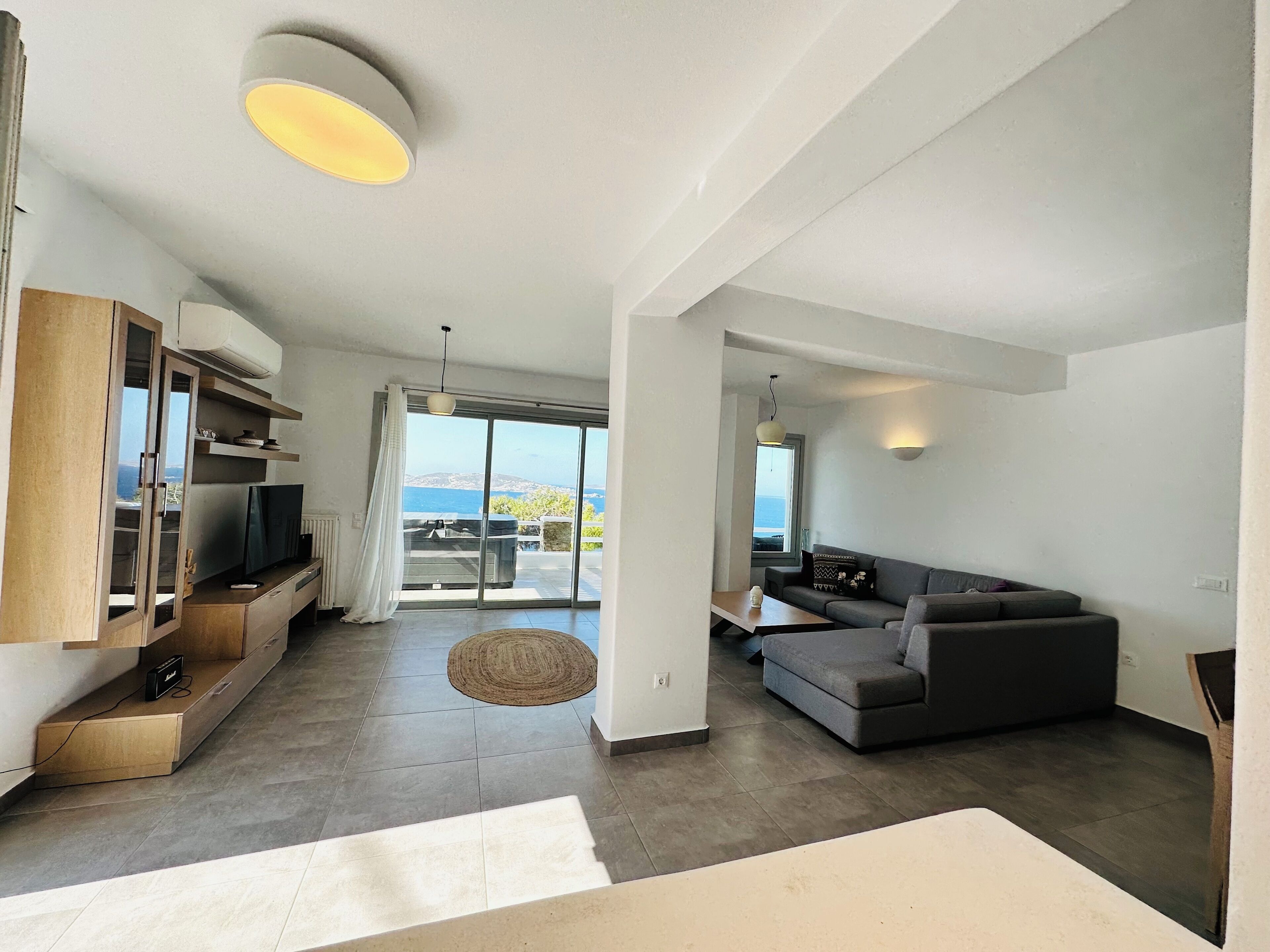 Uma sala de estar moderna com um sofá cinzento, uma televisão e uma janela ampla com vista para o mar.