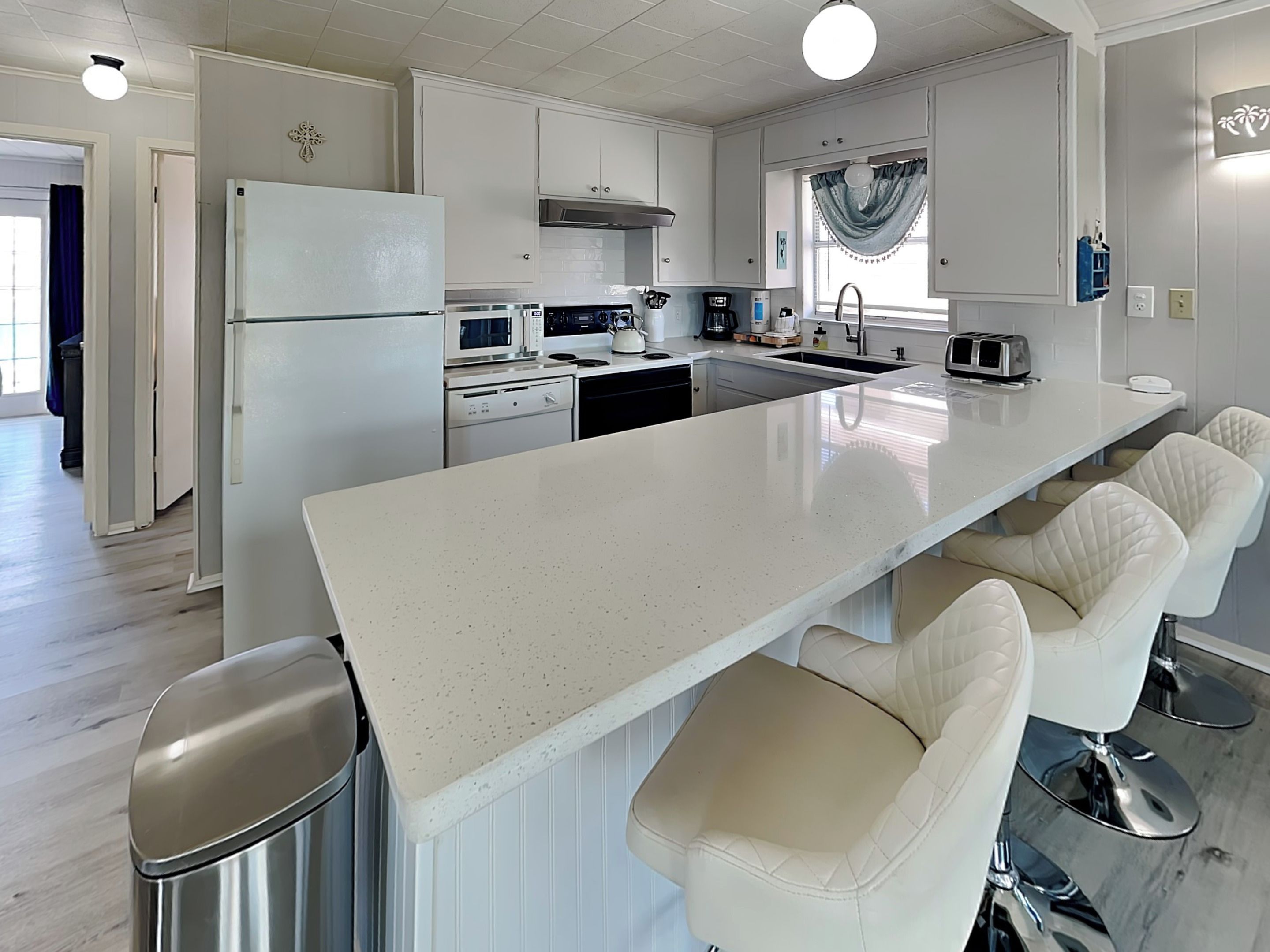 Uma cozinha moderna com armários brancos, uma ilha grande com banquetas, eletrodomésticos de aço inoxidável e uma janela com vista.