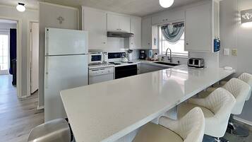 Uma cozinha moderna com armários brancos, uma ilha grande com banquetas, eletrodomésticos de aço inoxidável e uma janela com vista.