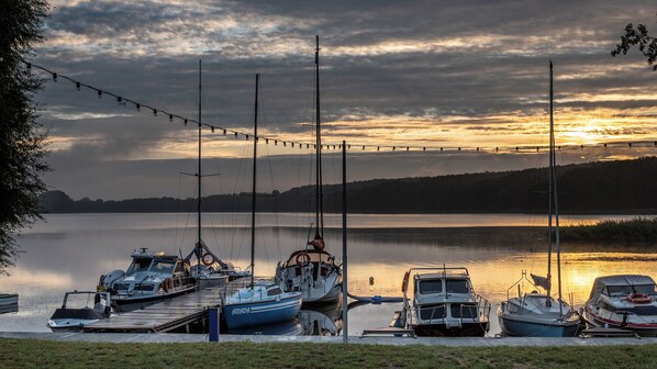 Marina com vários barcos atracados, uma ponte ao fundo e um pôr do sol sobre um lago calmo.