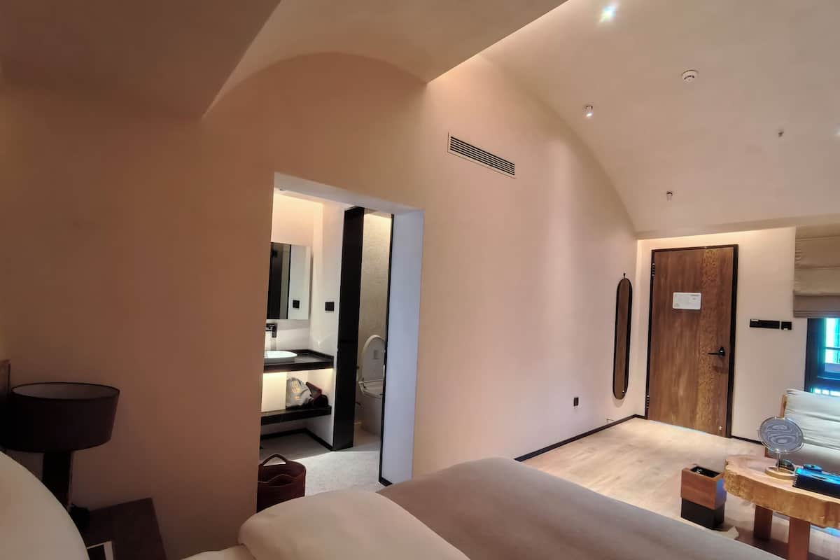 Quarto de hotel moderno com uma cama grande, uma mesa de cabeceira com abajur, uma porta de madeira e um banheiro visível através de uma porta aberta.