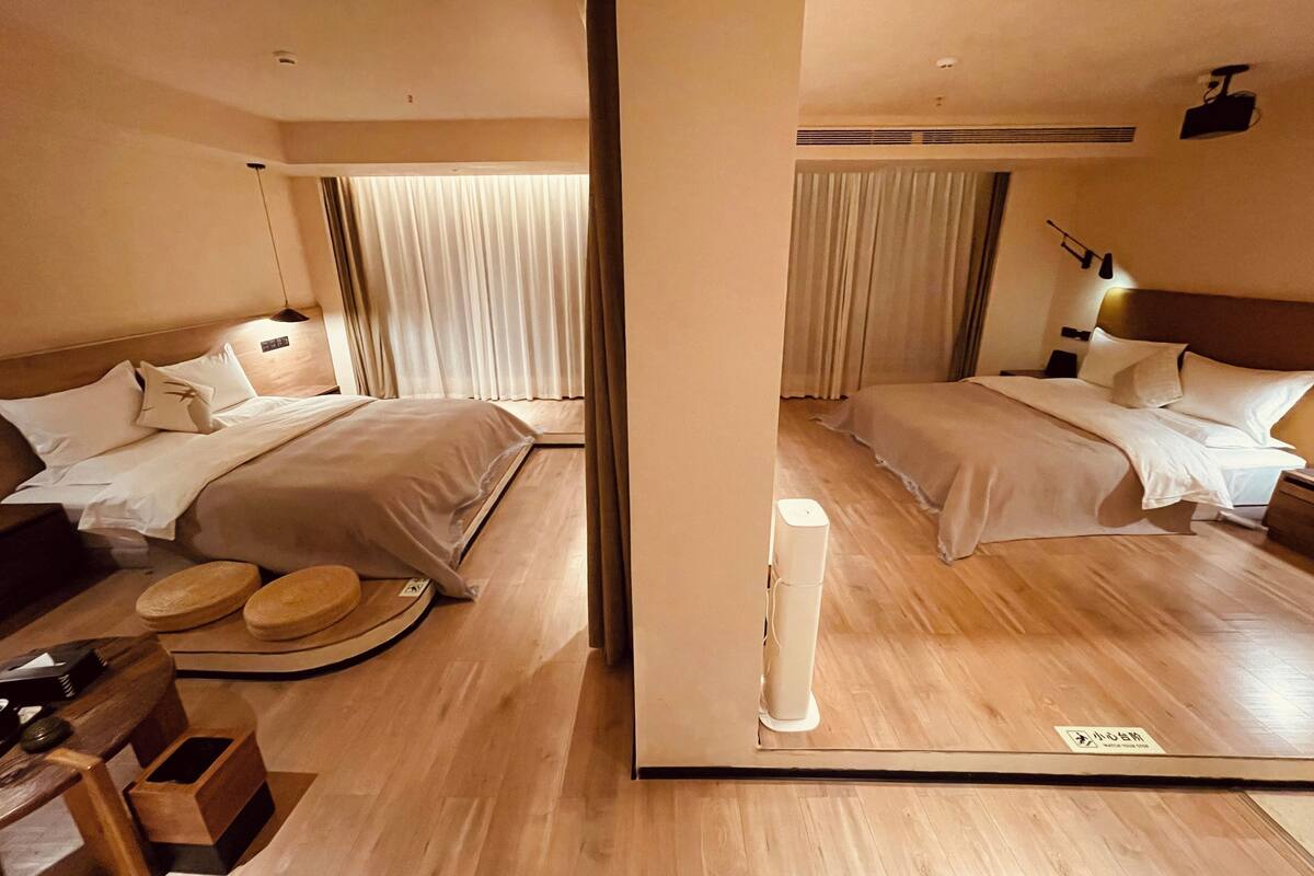 Quarto de hotel com duas camas, piso de madeira, um espelho grande e uma pequena mesa redonda com um chapéu.