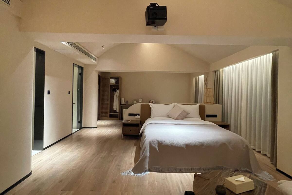 Quarto de hotel moderno com uma cama grande, uma mesa de cabeceira, uma mesinha com um telefone e um projetor no teto.