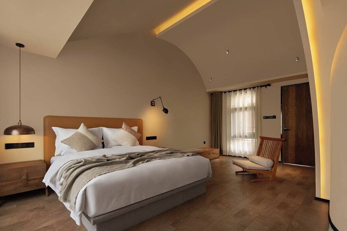 Quarto moderno com uma cama grande, uma mesa de cabeceira de madeira, um abajur e uma cadeira.