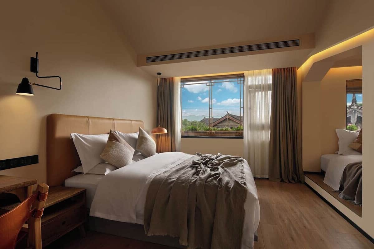 Quarto de hotel com cama, mesa de cabeceira, luminária, janela com cortinas e vista para arquitetura tradicional.