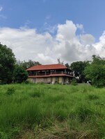 Uma casa de dois andares com telhado vermelho, rodeada por vegetação exuberante e árvores.