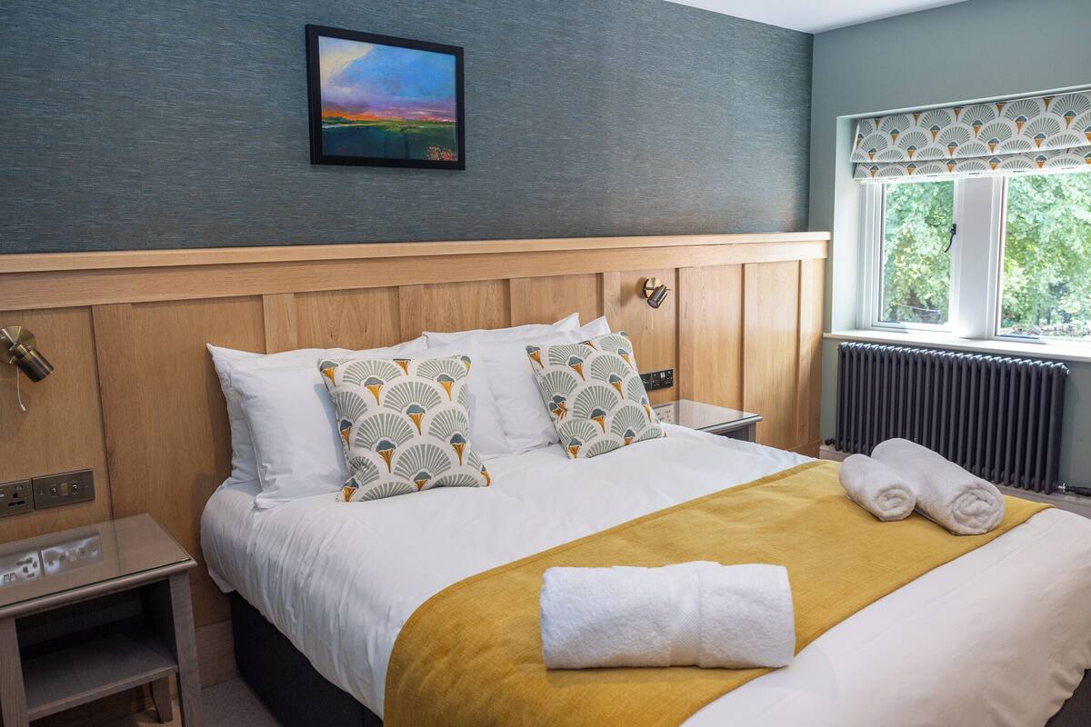 Cama bem arrumada com um cobertor amarelo, mesa de cabeceira com abajur e janela com vista para o verde.
