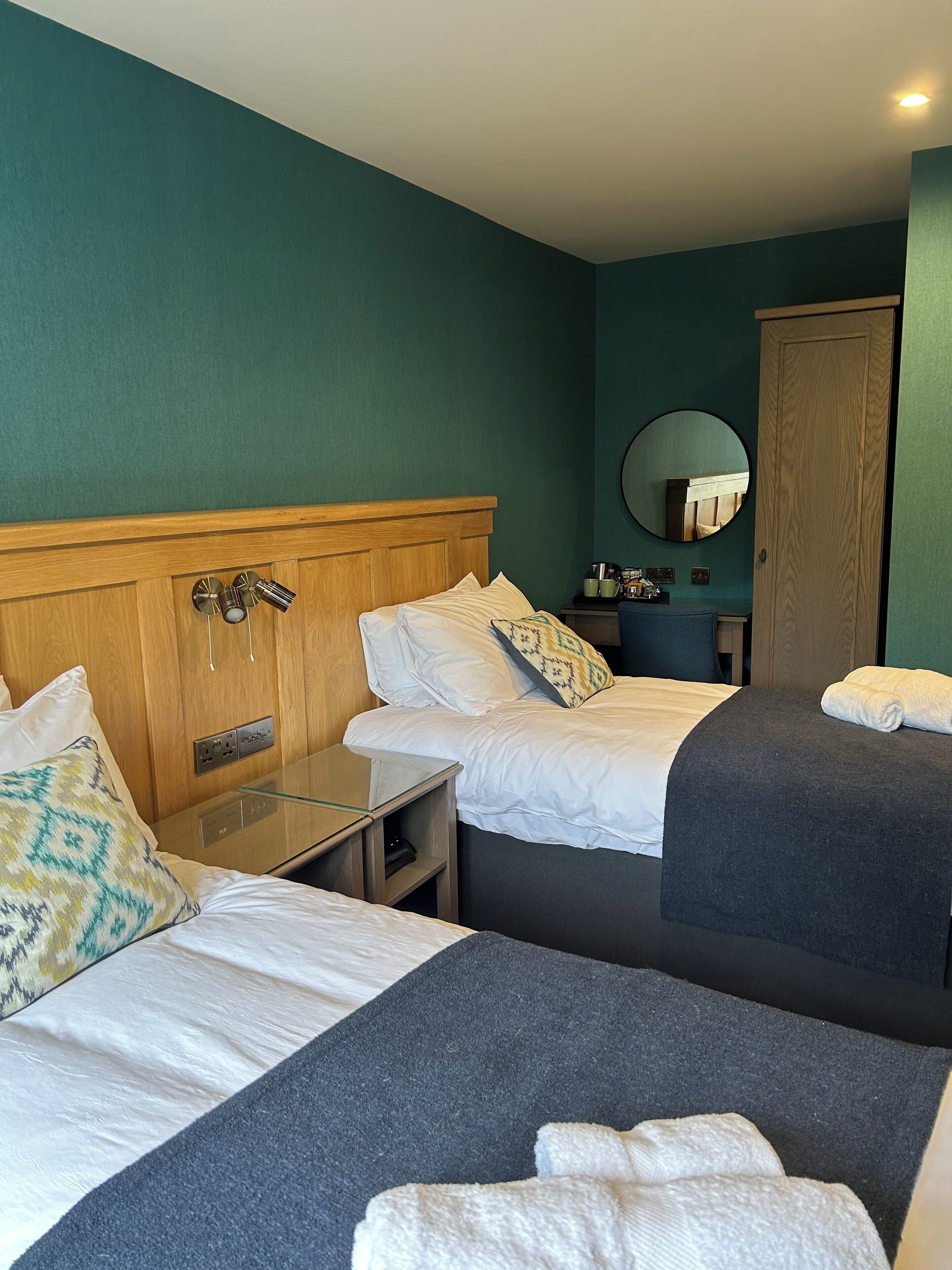 Quarto de hotel com duas camas, uma mesinha de cabeceira, um espelho e um guarda-roupa de madeira.