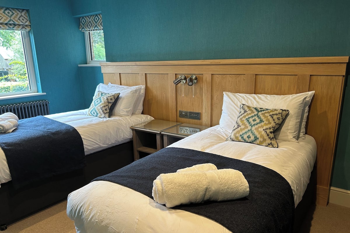 Quarto de hotel com duas camas, cada uma com lençóis brancos e almofadas estampadas, uma cabeceira de madeira, uma mesa de cabeceira com abajur e uma janela com cortinas estampadas.