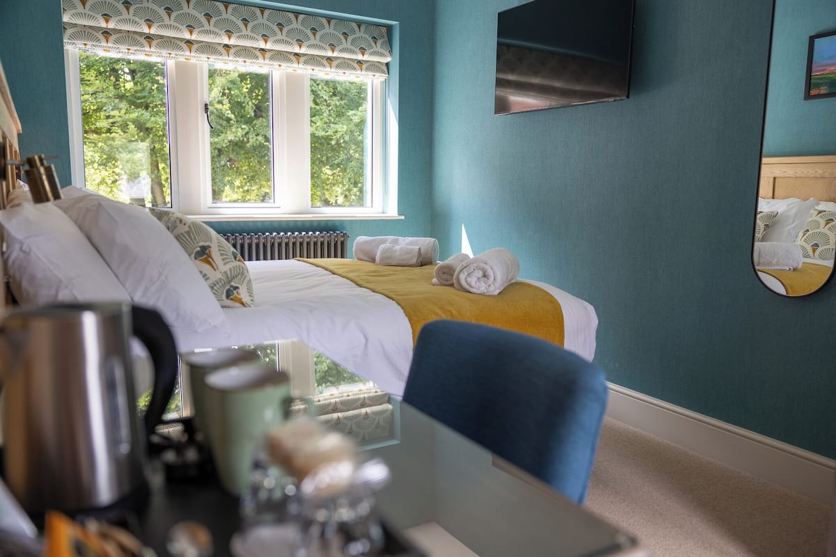Quarto de hotel com cama, mesa com chaleira e janela com vista para árvores.