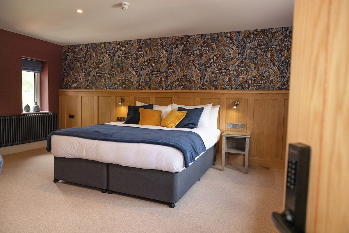 Quarto com uma cama grande, cabeceira de madeira, parede estampada, mesa de cabeceira e radiador.