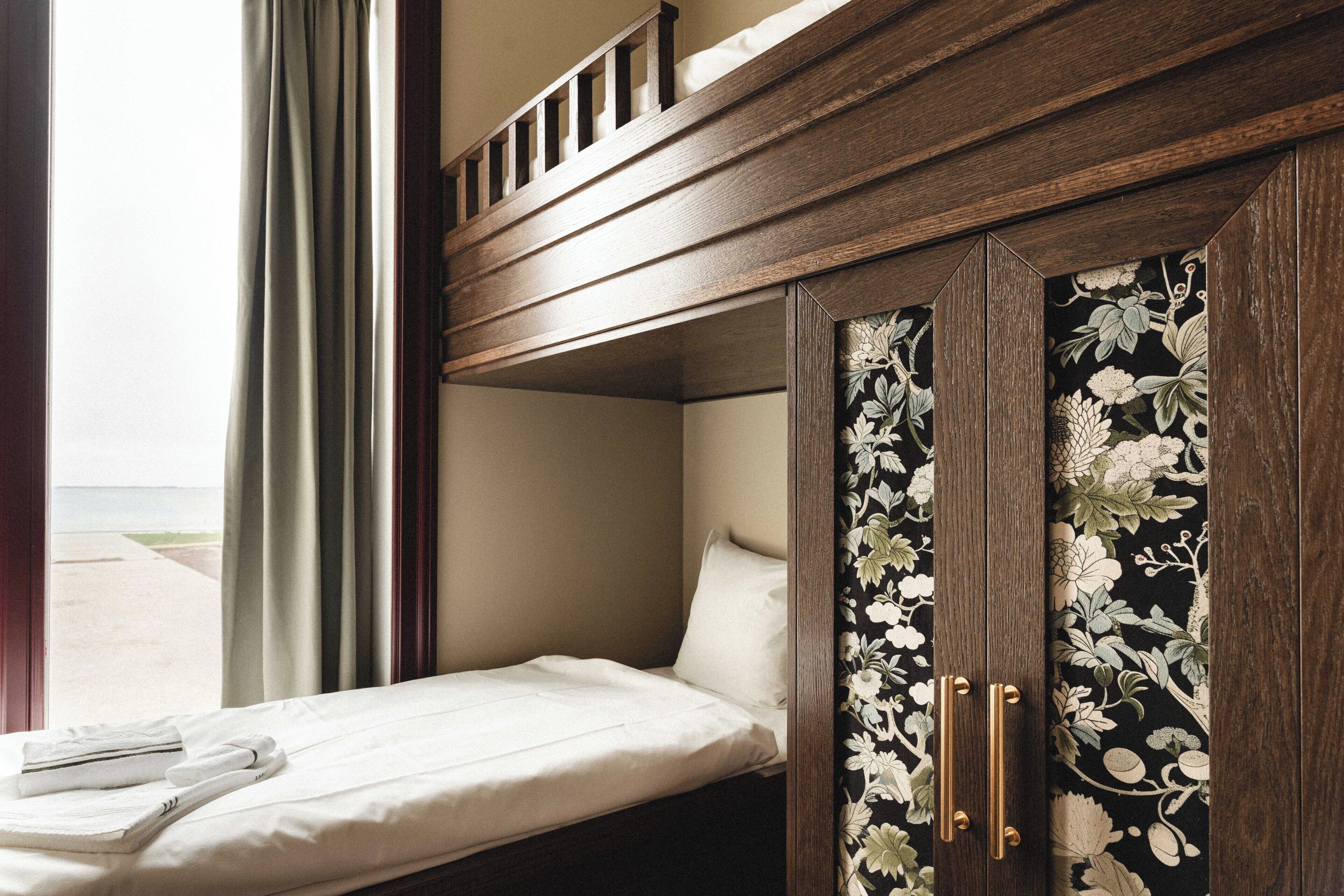 Uma cama de solteiro com cabeceira de madeira, uma janela com cortinas e um guarda-roupa com papel de parede floral.