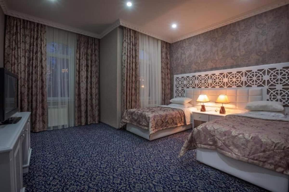 Quarto de hotel com duas camas, televisão, mesa de cabeceira com luminárias e tapete estampado.