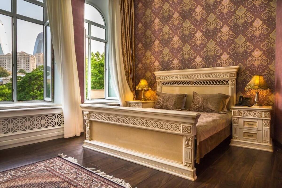 Quarto com uma cama grande, cabeceira ornamentada, duas mesas de cabeceira com abajures, parede estampada e vista de edifícios pela janela.