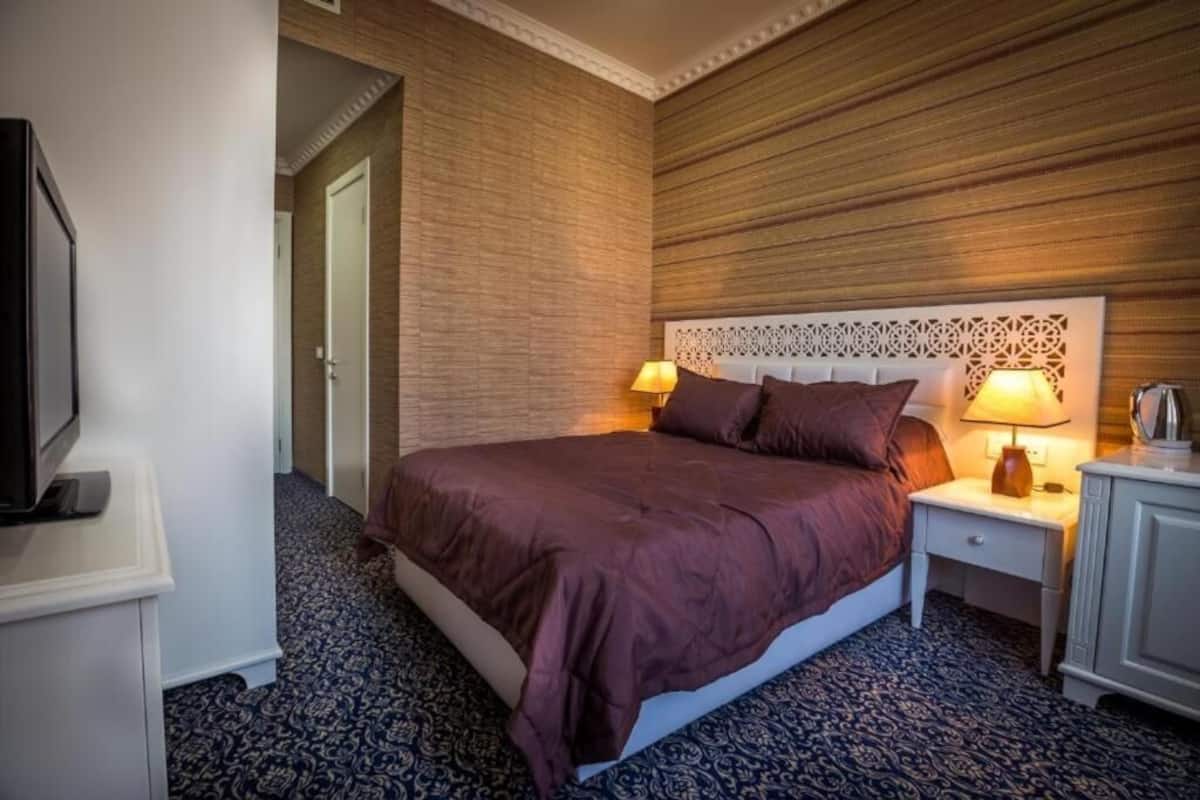 Quarto de hotel com uma cama grande, duas mesas de cabeceira com luminárias, uma televisão em um suporte e um tapete estampado.